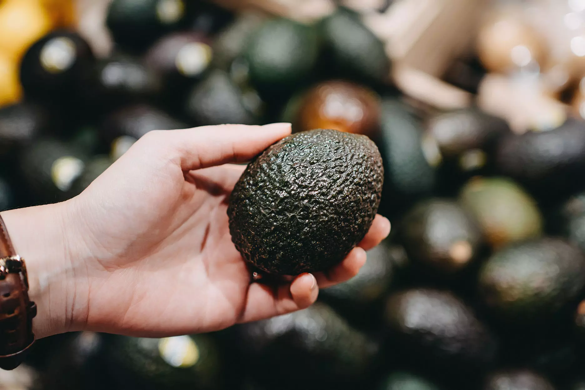 Bean ag siopadóireacht le haghaidh torthaí úra agus glasraí san ollmhargadh, gar di a lámh ag roghnú avocados