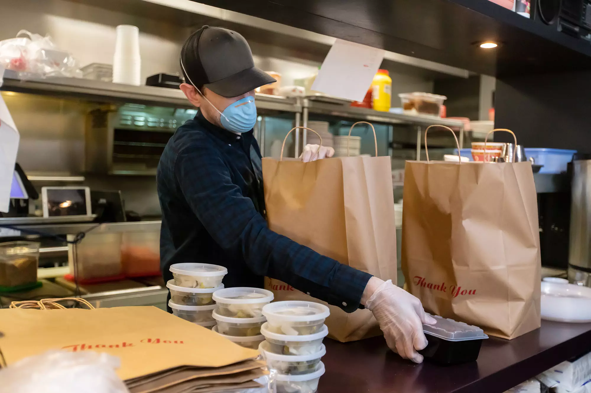 Mies hansikkaissa ja naamioissa ravintolakeittiössä valmistamassa ruokaa toimitettaviksi Covid-19-pandemian aikana