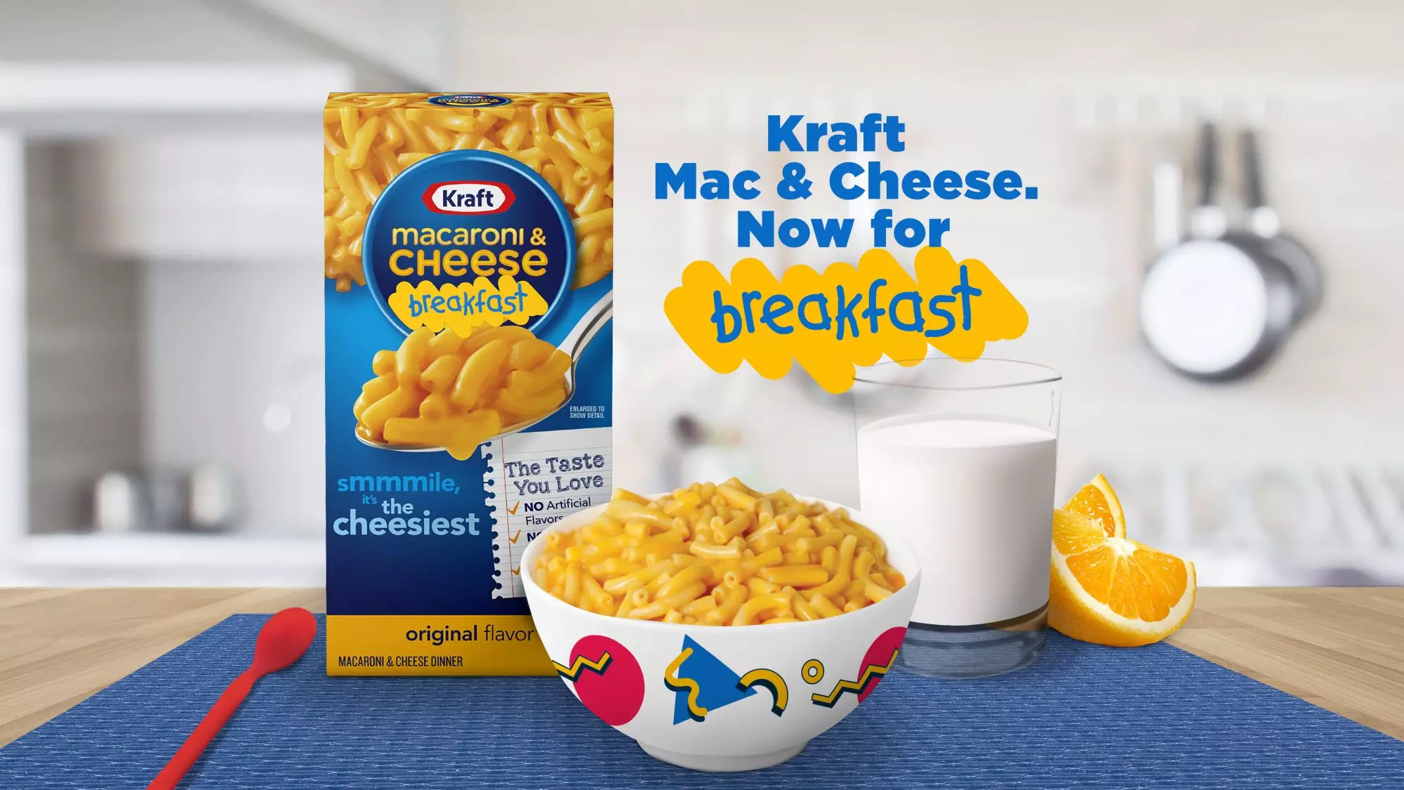 ប្រអប់ Kraft macaroni និងឈីសមួយដាក់នៅពីក្រោយចានប៉ាស្តាឈីសជាមួយទឹកដោះគោមួយកែវ និងចំណិតក្រូច